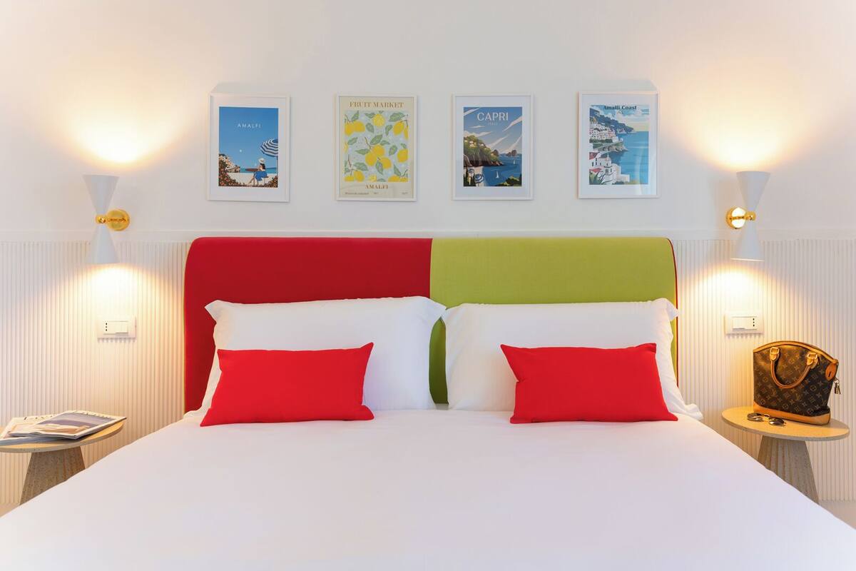 Quarto de hotel com cama, almofadas vermelhas e brancas, cabeceira vermelha e verde, e três quadros na parede.