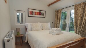 Quarto com cama de madeira, roupa de cama branca, radiador, pequena mesa com abajur e janela com cortinas.