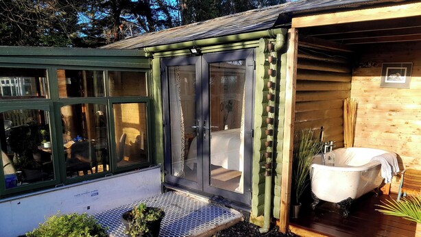 Uma cabana de madeira com uma janela grande, uma porta para um banheiro com banheira e uma pequena varanda com plantas em vasos.