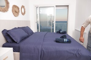 Quarto com cama, cômoda, vista do mar através de uma porta deslizante e uma pessoa parada perto da porta.