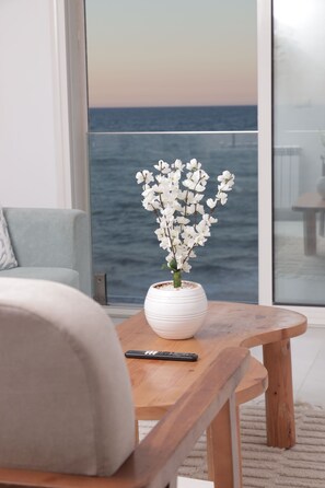 Mesa de madeira com um vaso branco contendo flores brancas, um controle remoto e vista do oceano por uma porta de vidro.