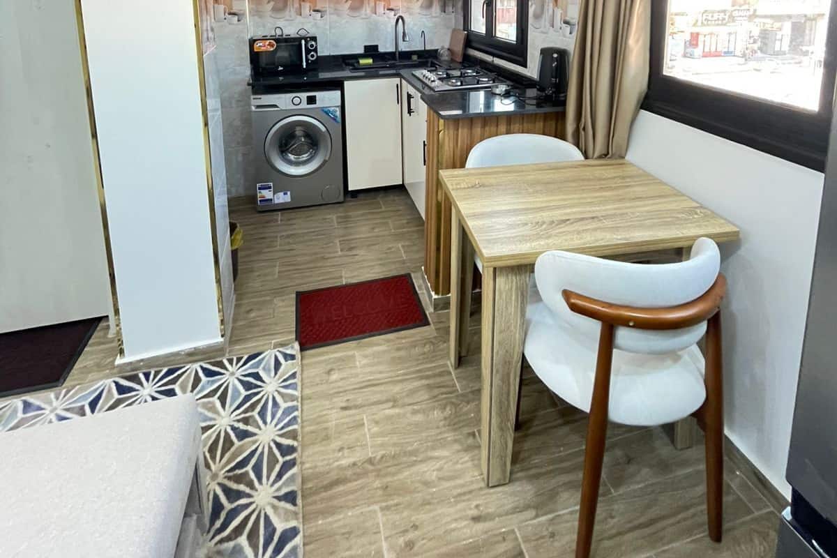 Cozinha compacta com máquina de lavar, área de refeições com mesa e cadeiras, e janela com cortinas.