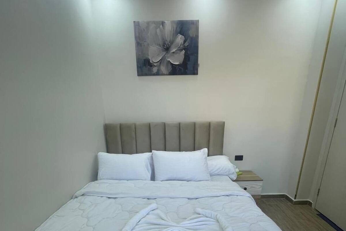 Uma cama bem arrumada com cabeceira, um quadro acima dela e uma mesinha de cabeceira de madeira com uma luminária ao lado.