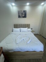 Uma cama bem arrumada com cabeceira, um quadro acima dela e uma mesinha de cabeceira de madeira com uma luminária ao lado.