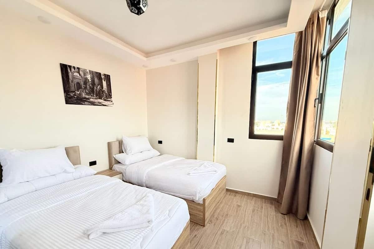 Quarto de hotel com duas camas, uma janela grande com cortinas, um quadro na parede e chão de cor clara.