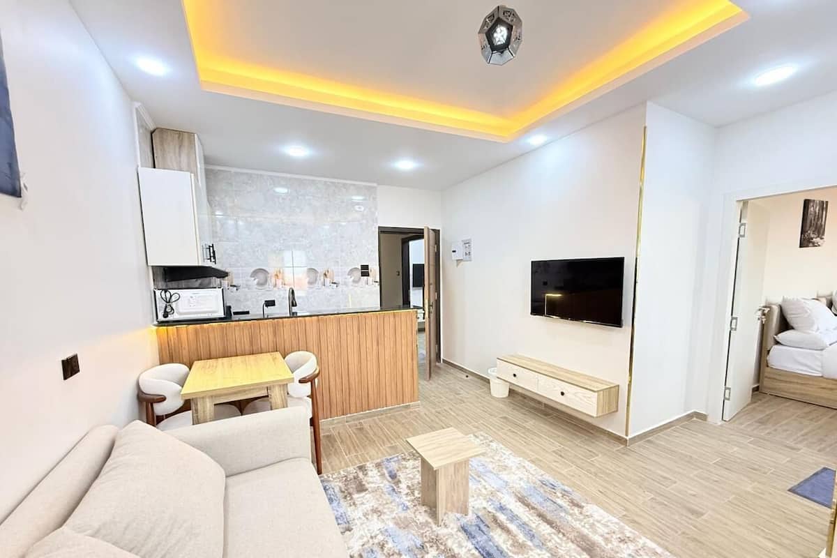 Uma sala de estar moderna com um sofá, uma mesa de centro, uma televisão e uma cozinha ao fundo.