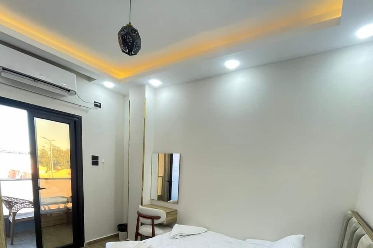 Quarto moderno com cama grande, cabeceira de madeira, mesa de cabeceira pequena, uma cadeira, espelho e porta de vidro deslizante para área externa.