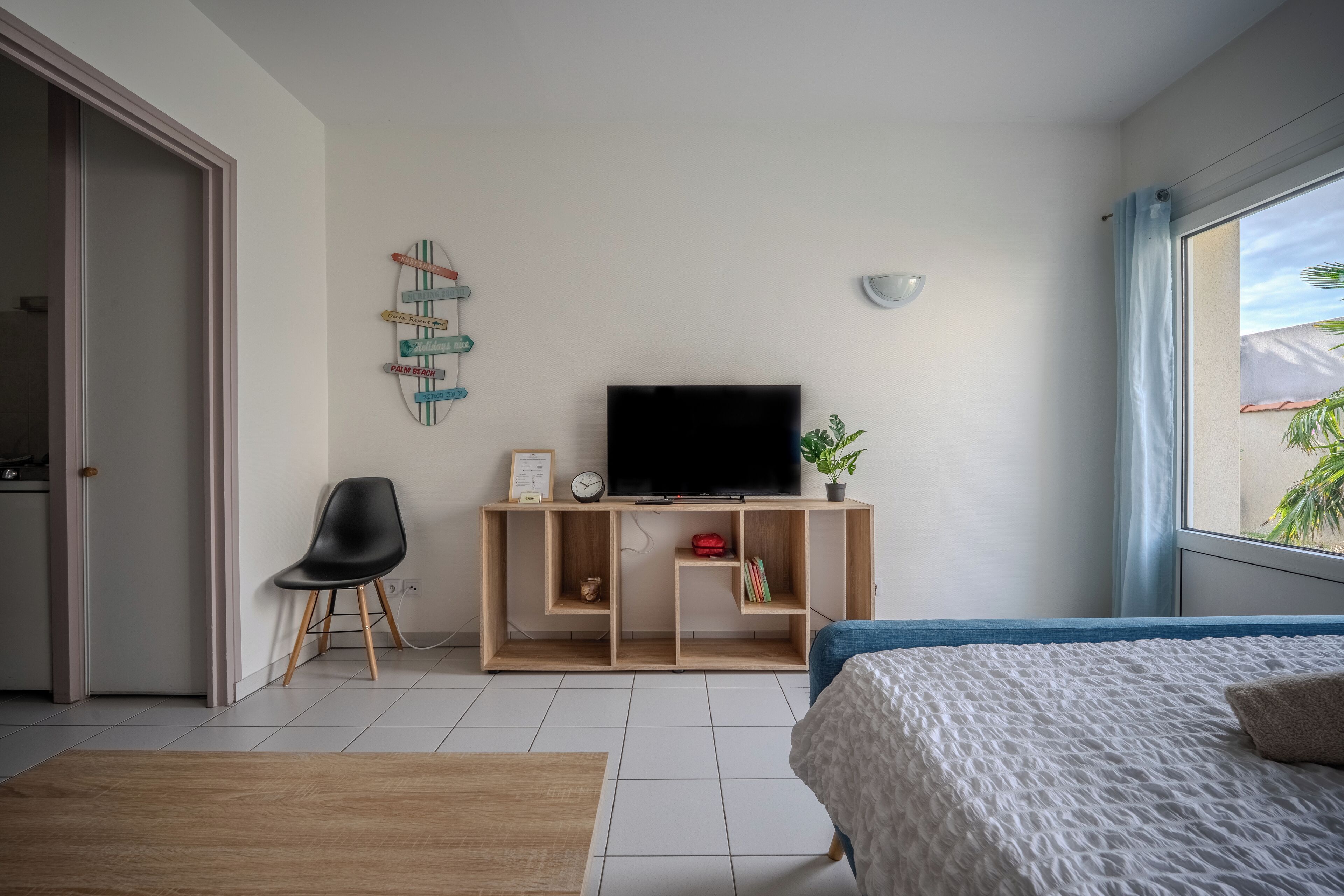 Quarto de hotel moderno com uma cama, uma televisão sobre um suporte de madeira, uma cadeira, uma pequena mesa e uma janela com cortinas.