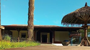 Um edifício com entrada coberta, ampla área externa com assentos e um telhado de palha, e um caminho pavimentado que leva à entrada.