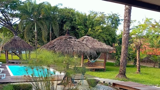 Uma área de piscina com um elemento aquático, rodeada por vegetação tropical e estruturas com telhado de palha.