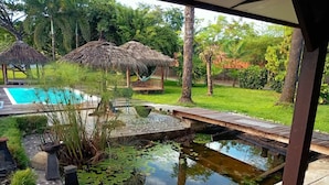 Uma área de piscina ao ar livre tranquila, com um pequeno lago, rodeada por vegetação exuberante e uma estrutura com telhado de palha.