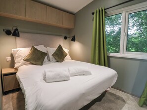 Cama bem arrumada com lençóis brancos e duas almofadas, mesa de cabeceira com abajur, janela com cortinas verdes e cabeceira de madeira.
