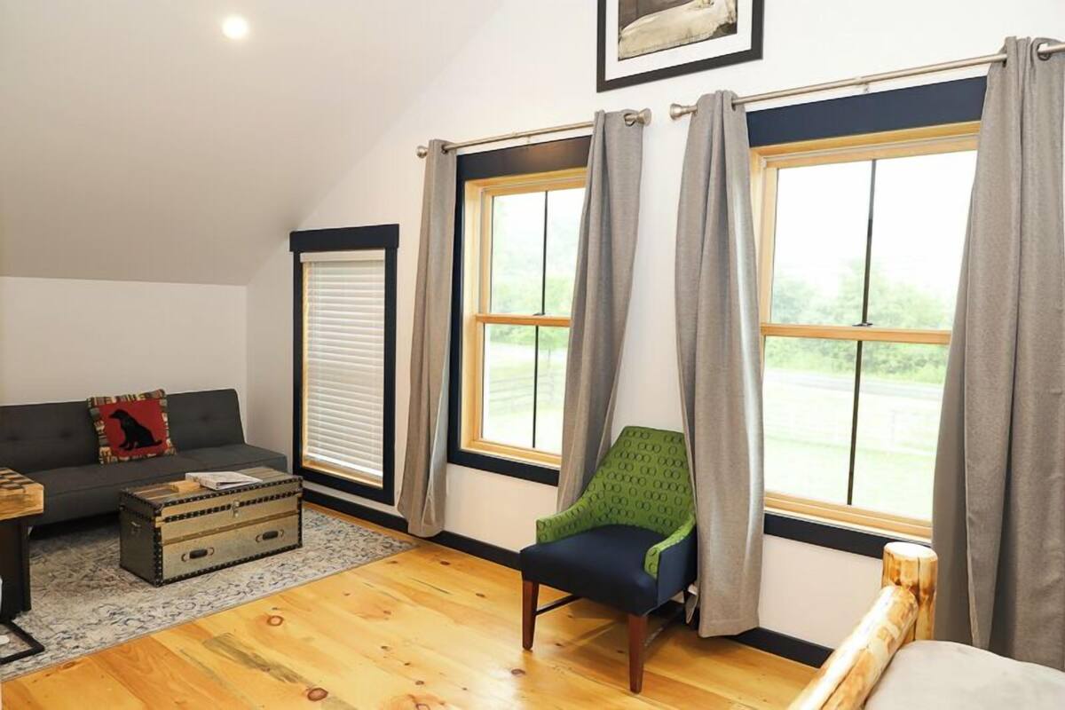 Um quarto com piso de madeira, uma cadeira verde, um sofá cinza, uma mesa de madeira e uma janela com cortinas.