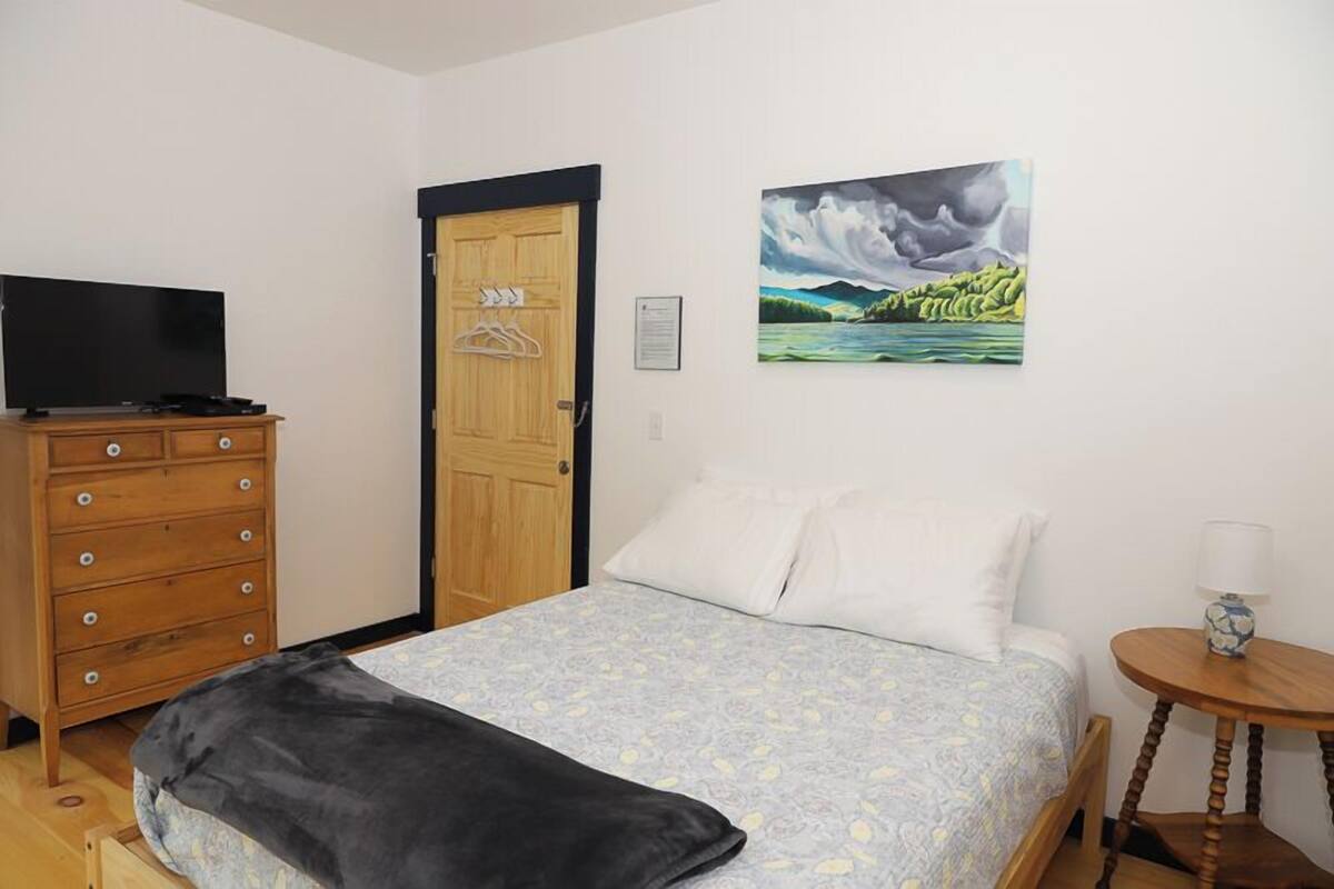 Quarto com cama, cômoda, televisão, pintura e mesinha com abajur.