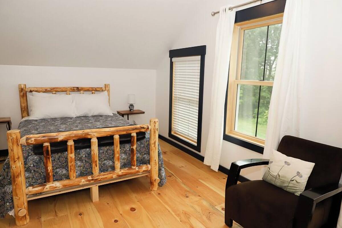 Quarto com cama de madeira, mesa de cabeceira, uma cadeira, janela com cortinas e vista para o verde lá fora.