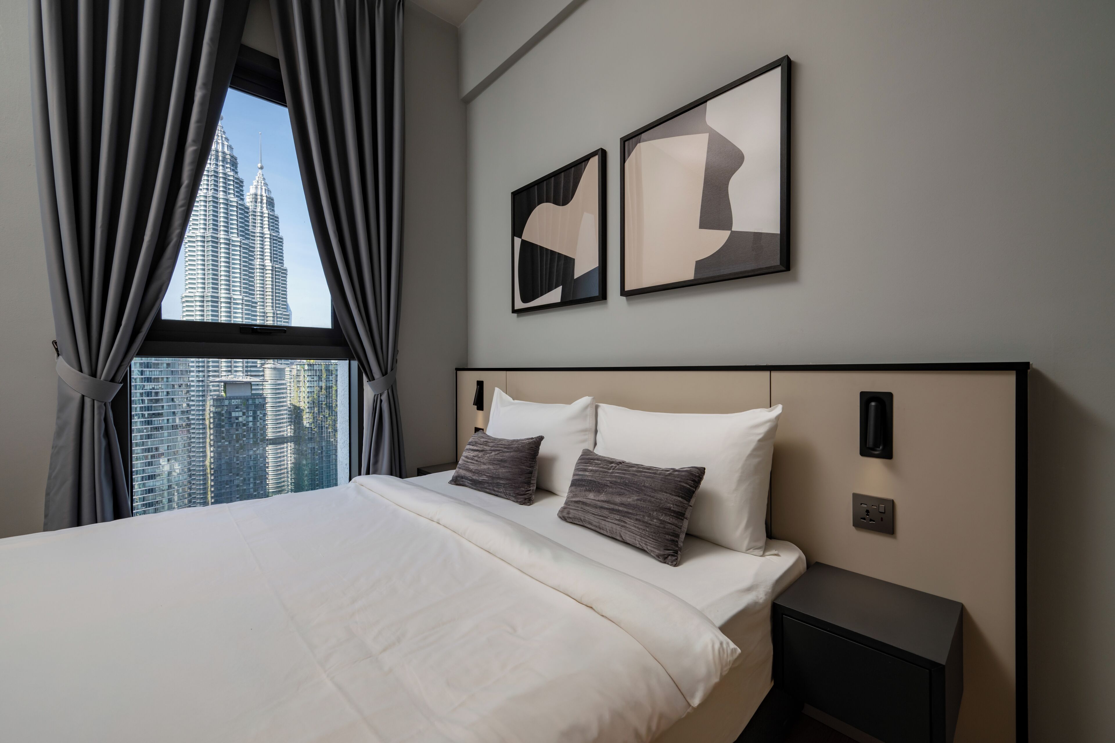 Quarto de hotel com uma cama grande, duas almofadas, uma mesa de cabeceira e uma janela com vista para a paisagem urbana.