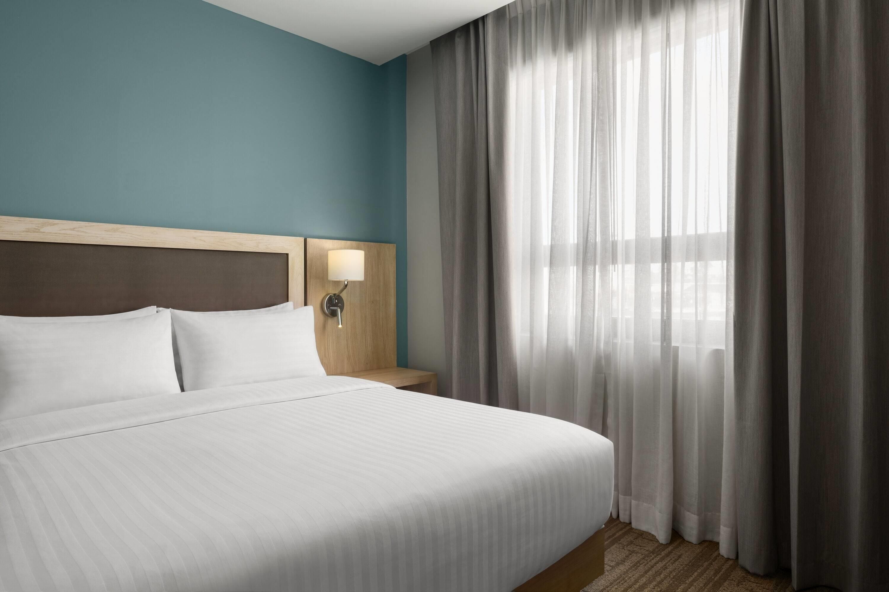 Quarto de hotel com cama, mesa de cabeceira, luminária e cortinas finas.