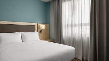 Quarto de hotel com cama, mesa de cabeceira, luminária e cortinas finas.