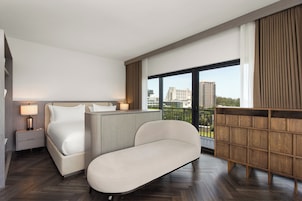 Quarto de hotel moderno com uma cama, um sofá, uma mesa de cabeceira e vista da cidade por grandes janelas.