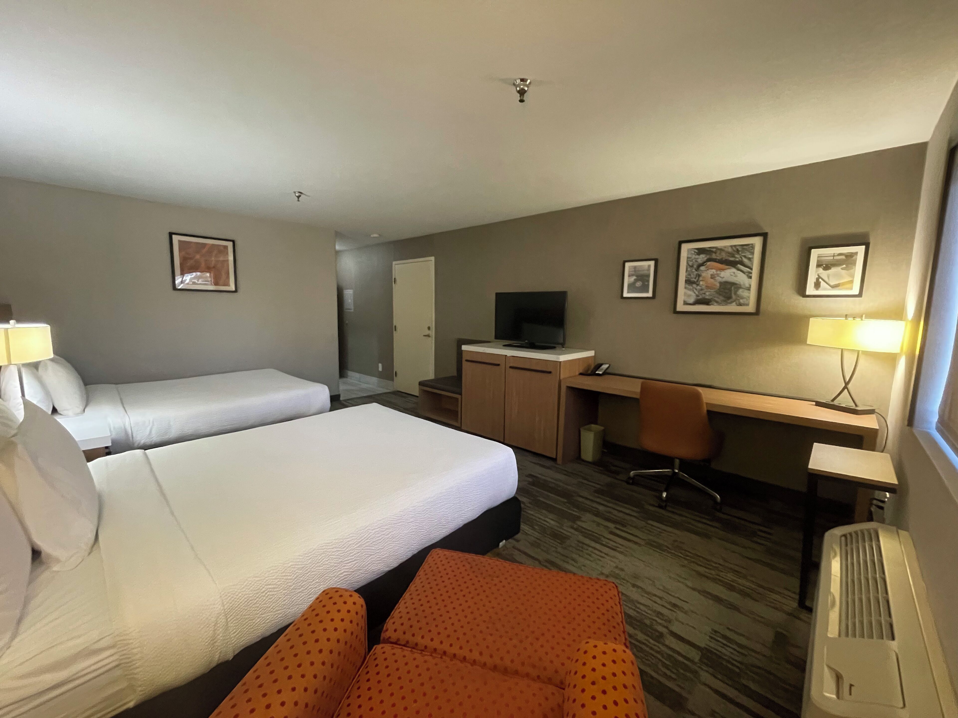 Quarto de hotel com duas camas de solteiro, uma escrivaninha com computador, televisão e um pequeno sofá laranja.