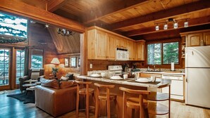Uma cozinha acolhedora em estilo cabana, com armários de madeira, uma ilha central com banquetas e uma área de refeições com sofá e cadeiras.