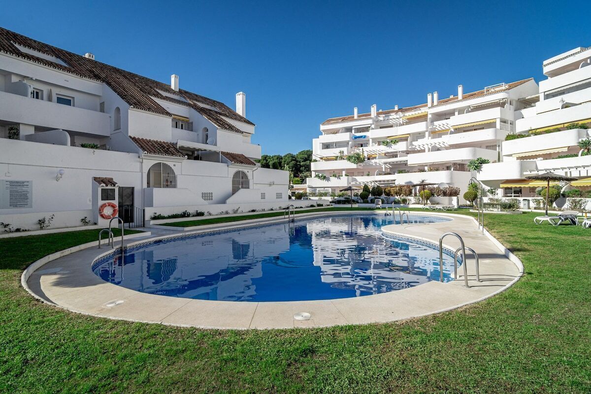 Uma piscina rodeada por um espaço gramado, com edifícios brancos com varandas e um telhado em azulejos ao fundo.
