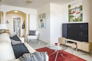 Uma sala de estar moderna com um sofá, uma mesa de centro de vidro, uma TV em um suporte de madeira e uma pintura de flamingos.
