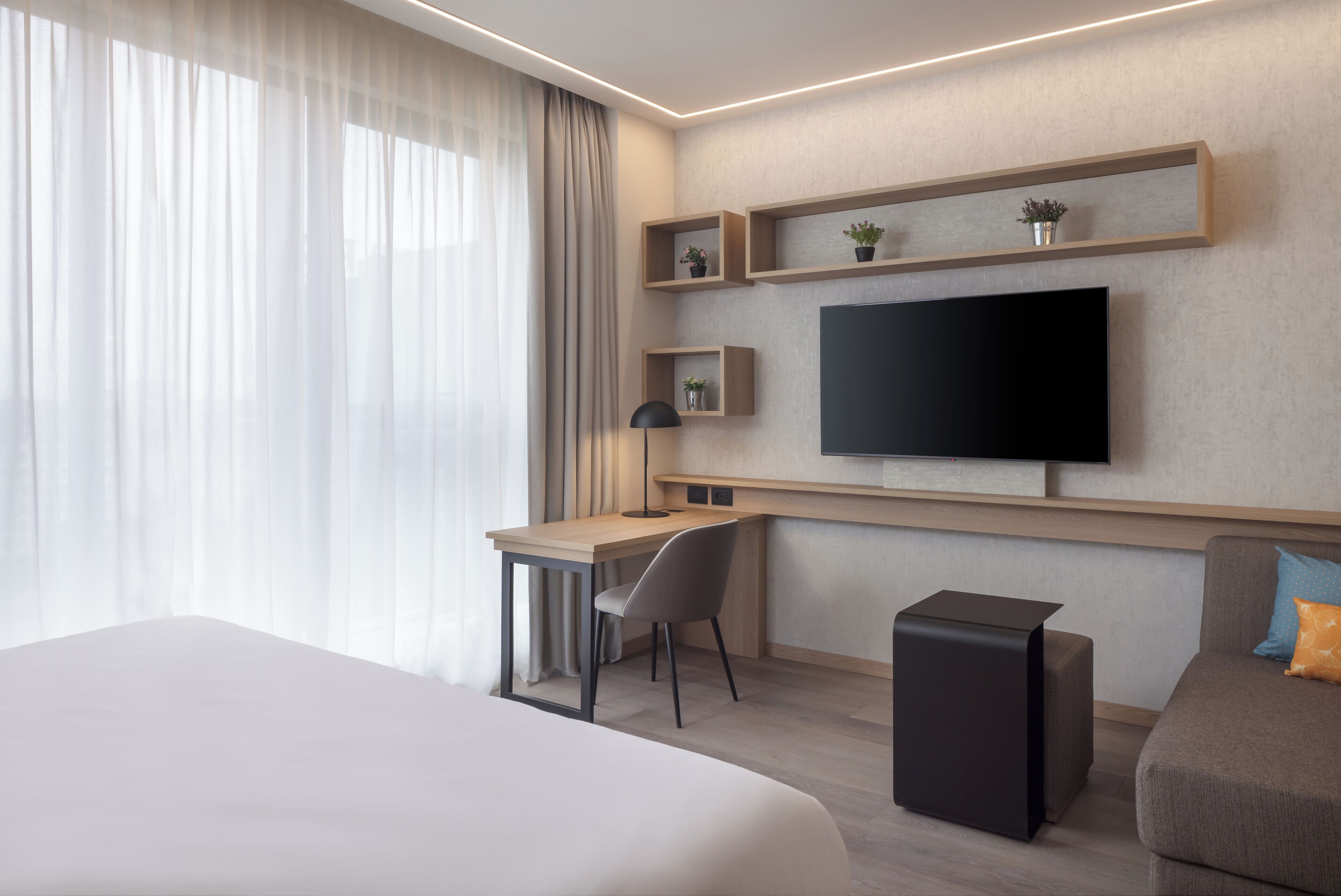 Quarto de hotel moderno com uma cama grande, uma escrivaninha com cadeira, televisão na parede e um espaço de estar pequeno com sofá e almofadas.