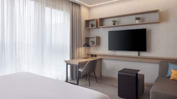 Quarto de hotel moderno com uma cama grande, uma escrivaninha com cadeira, televisão na parede e um espaço de estar pequeno com sofá e almofadas.