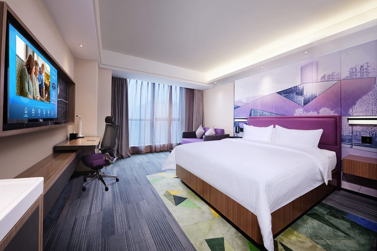 Quarto de hotel moderno com uma cama grande, uma escrivaninha com computador, uma TV na parede e uma janela ampla com cortinas.
