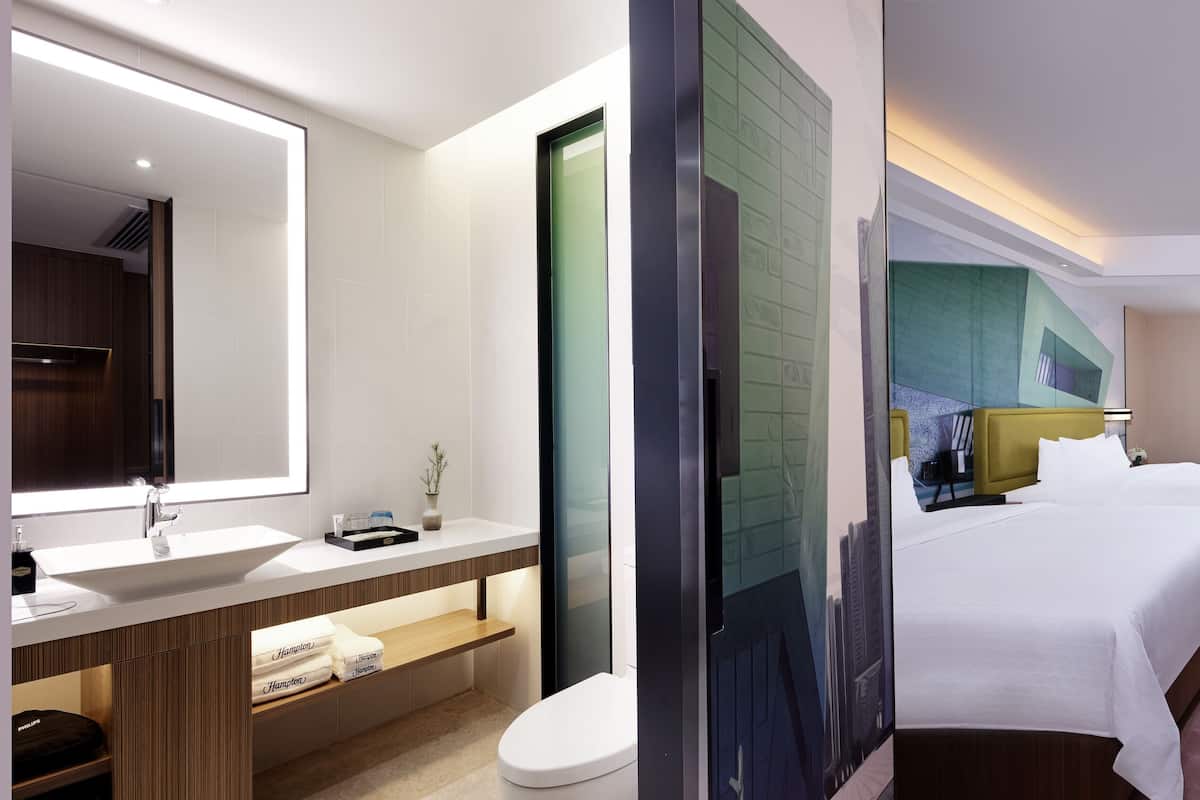 Quarto de hotel com uma cama grande, uma escrivaninha com cadeira, televisão, banheiro com pia e espelho, e janela com cortinas.