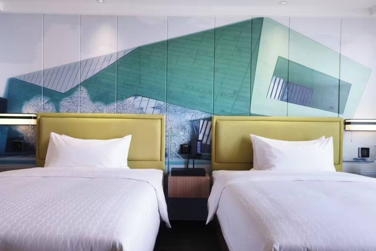 Quarto de hotel com duas camas, um mural moderno na parede e uma pequena mesa com abajur.