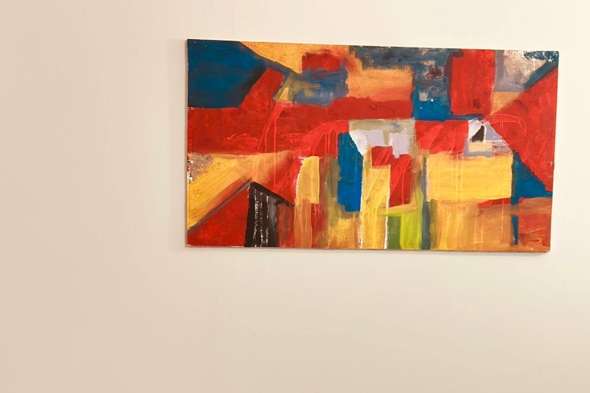 Quarto de hotel com cama, almofadas brancas, colcha azul e um quadro abstrato colorido na parede.