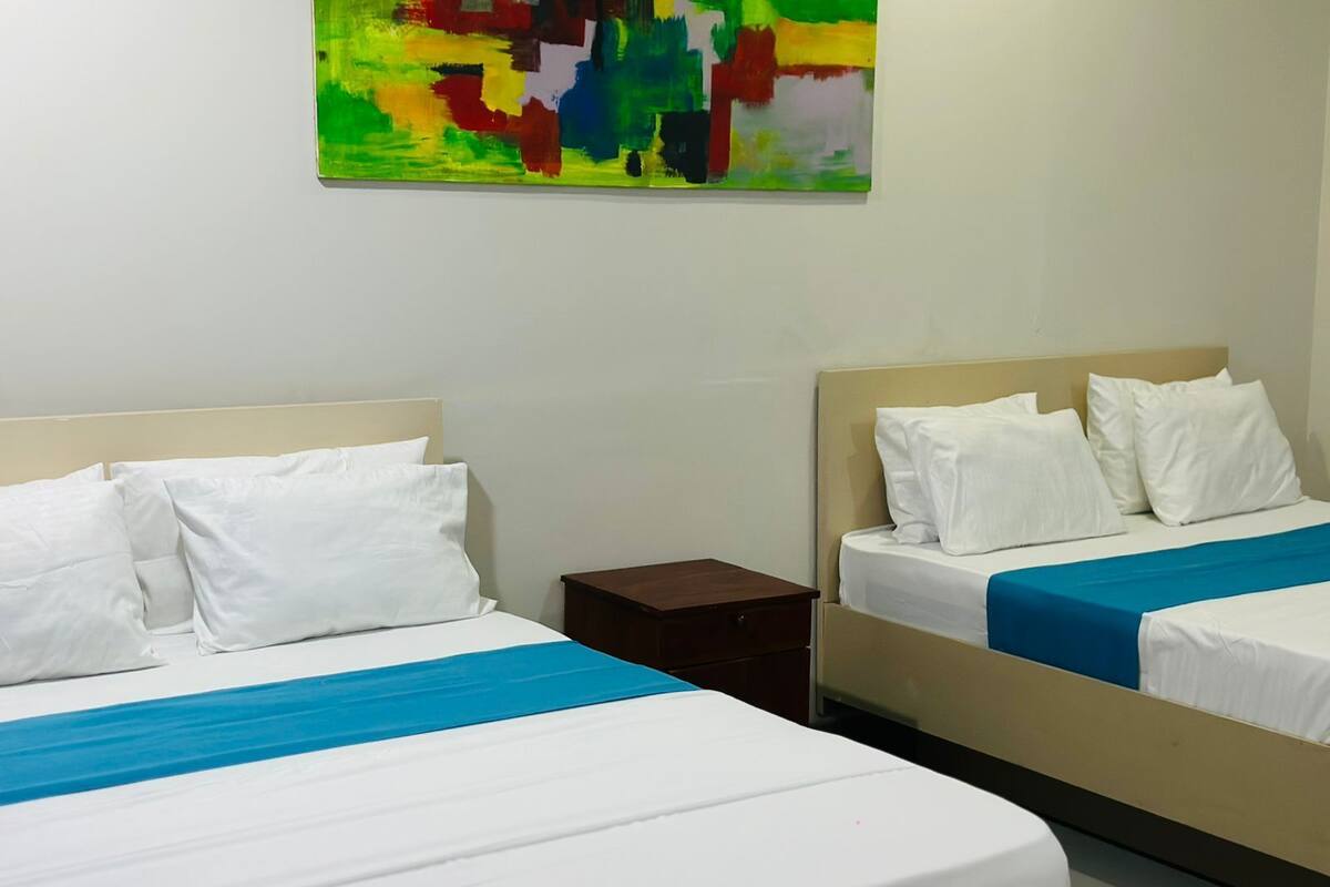 Quarto de hotel com duas camas, cada uma com roupa de cama branca e detalhes em azul, uma pequena mesa de cabeceira de madeira e um quadro abstrato colorido na parede.