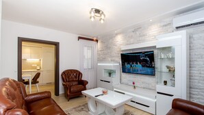 Uma sala de estar moderna com sofás de couro, uma mesa de centro branca, uma TV fixada em uma parede de pedra e um aparador de vidro com objetos decorativos.