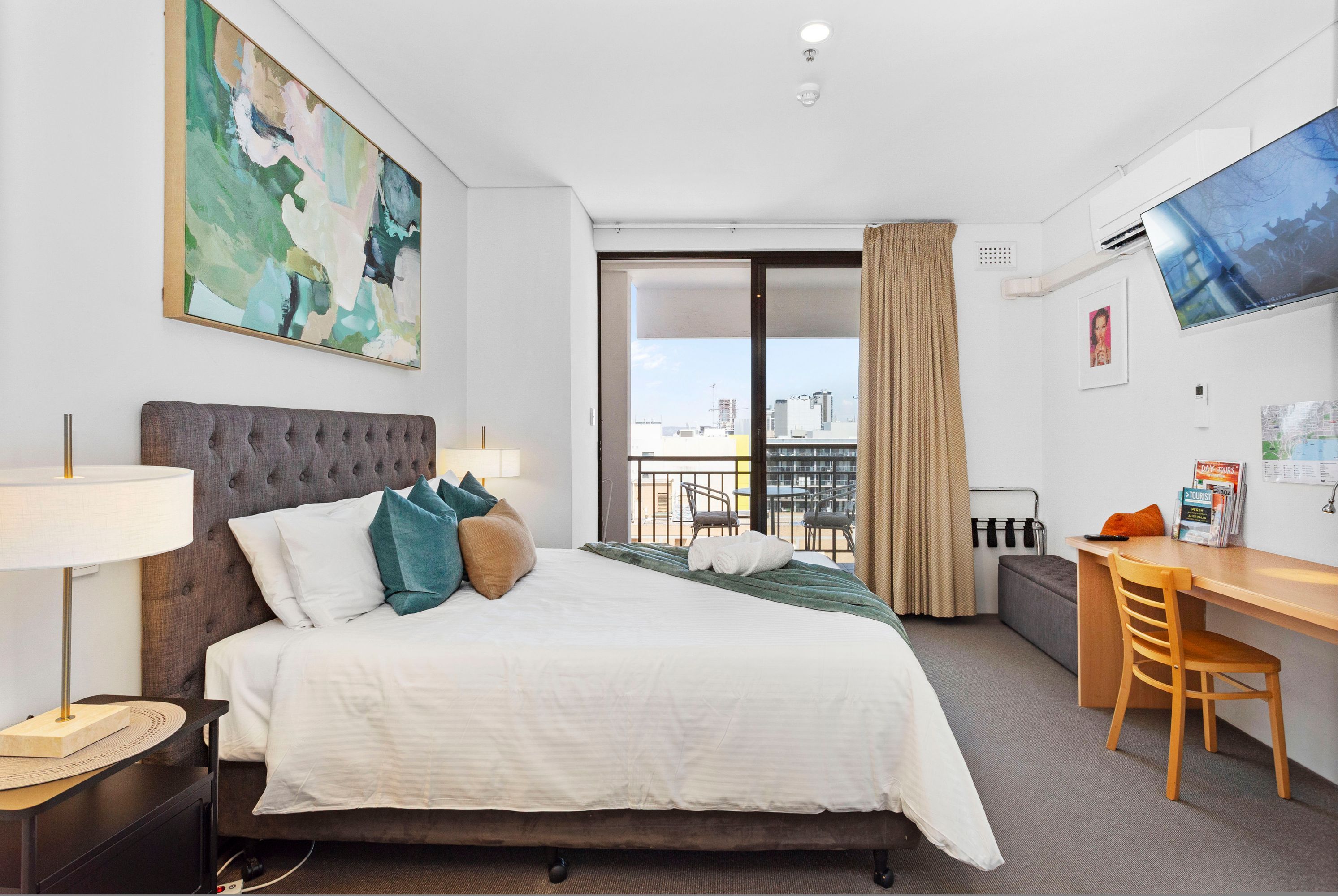 Quarto de hotel com cama, escrivaninha, cadeira, televisão e varanda com vista para a cidade.