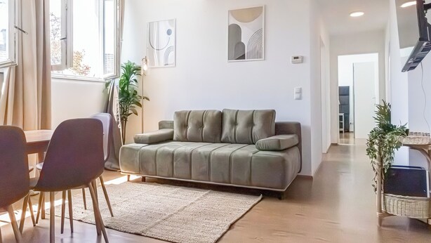 Uma sala de estar moderna com um sofá cinza, uma mesa de jantar com cadeiras, uma janela grande e um lustre.