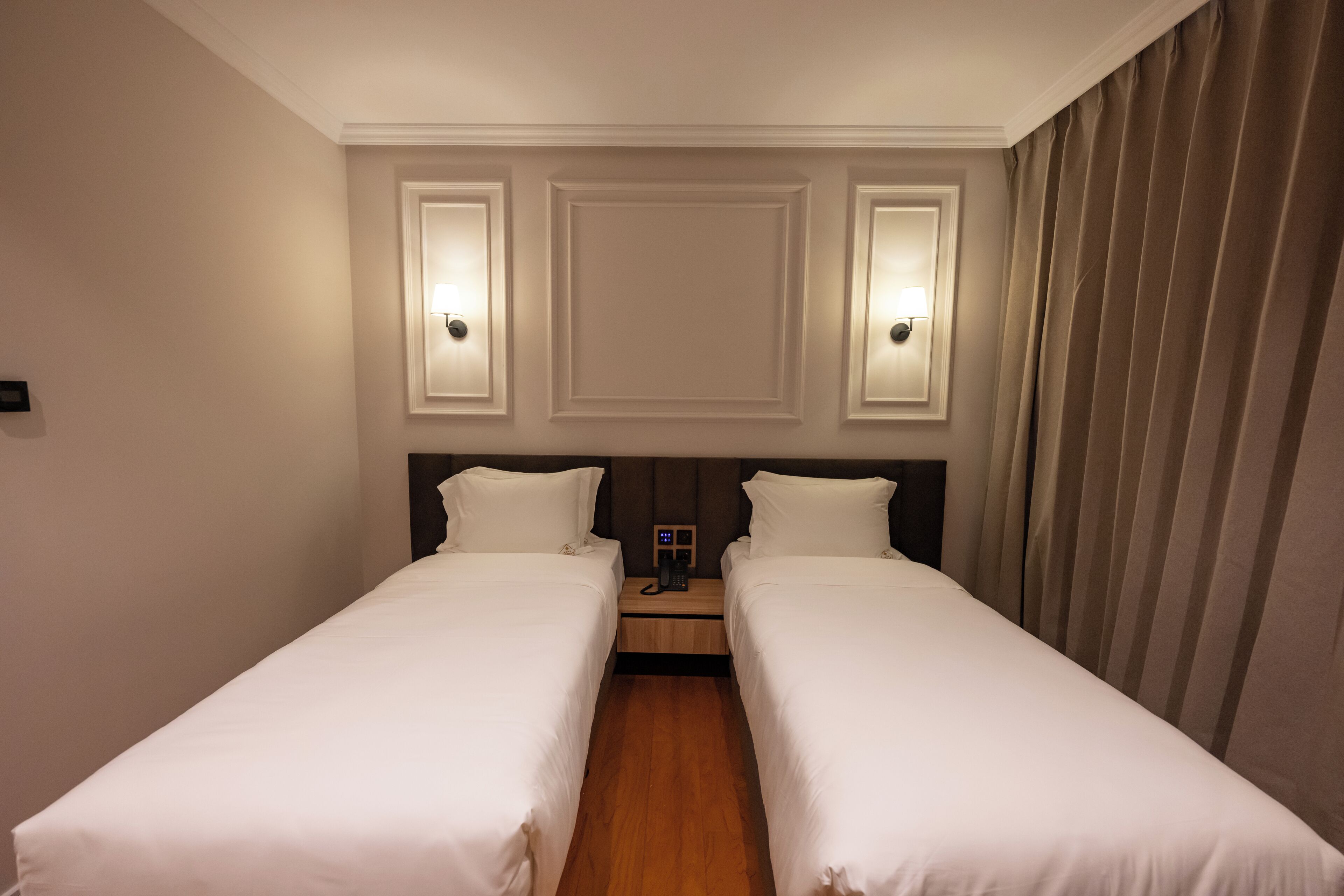 Quarto de hotel com duas camas de solteiro, uma mesa de cabeceira com abajur e luminária na parede.