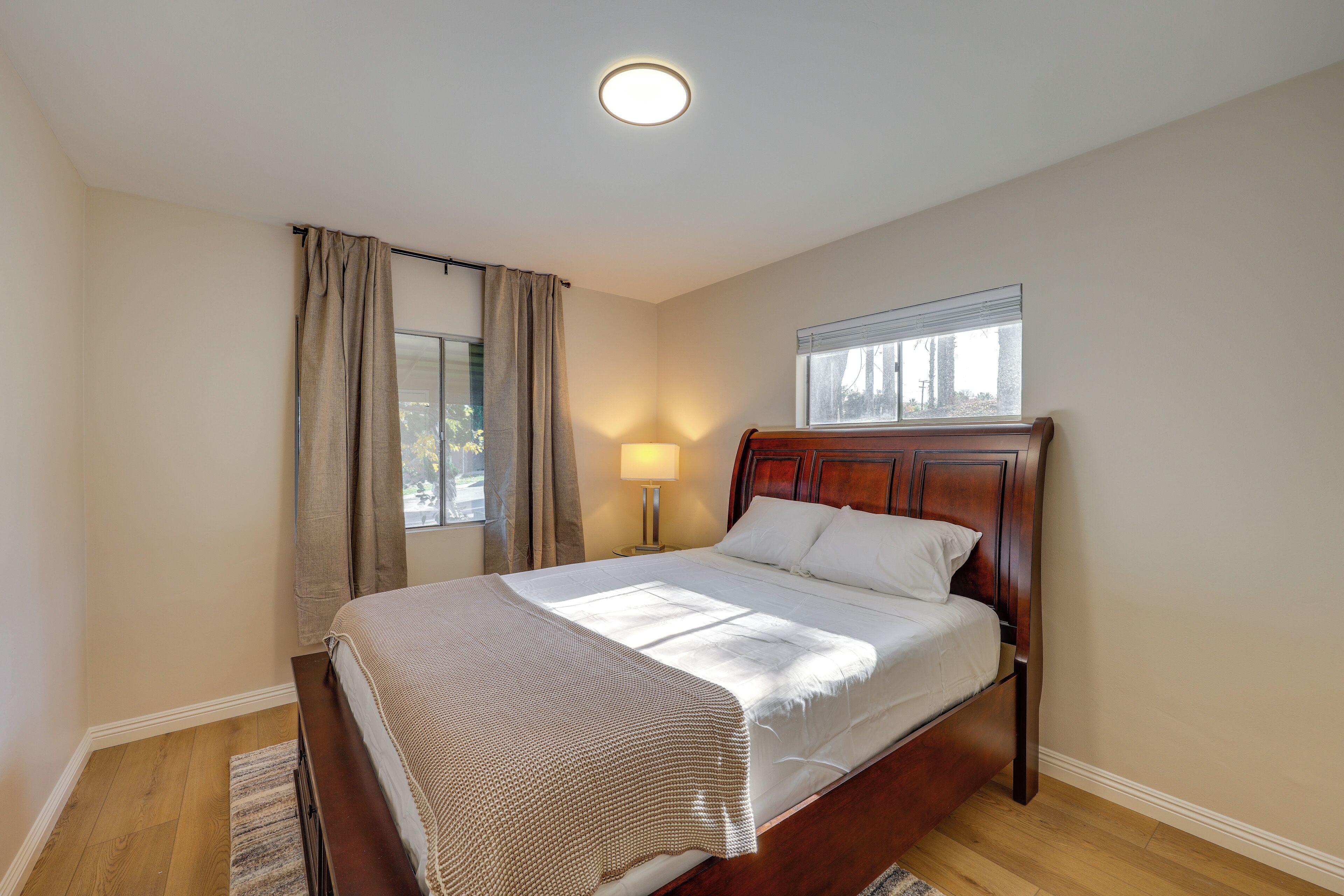 Quarto com cama de madeira, luminária de cabeceira, janela com cortinas e vista para árvores.