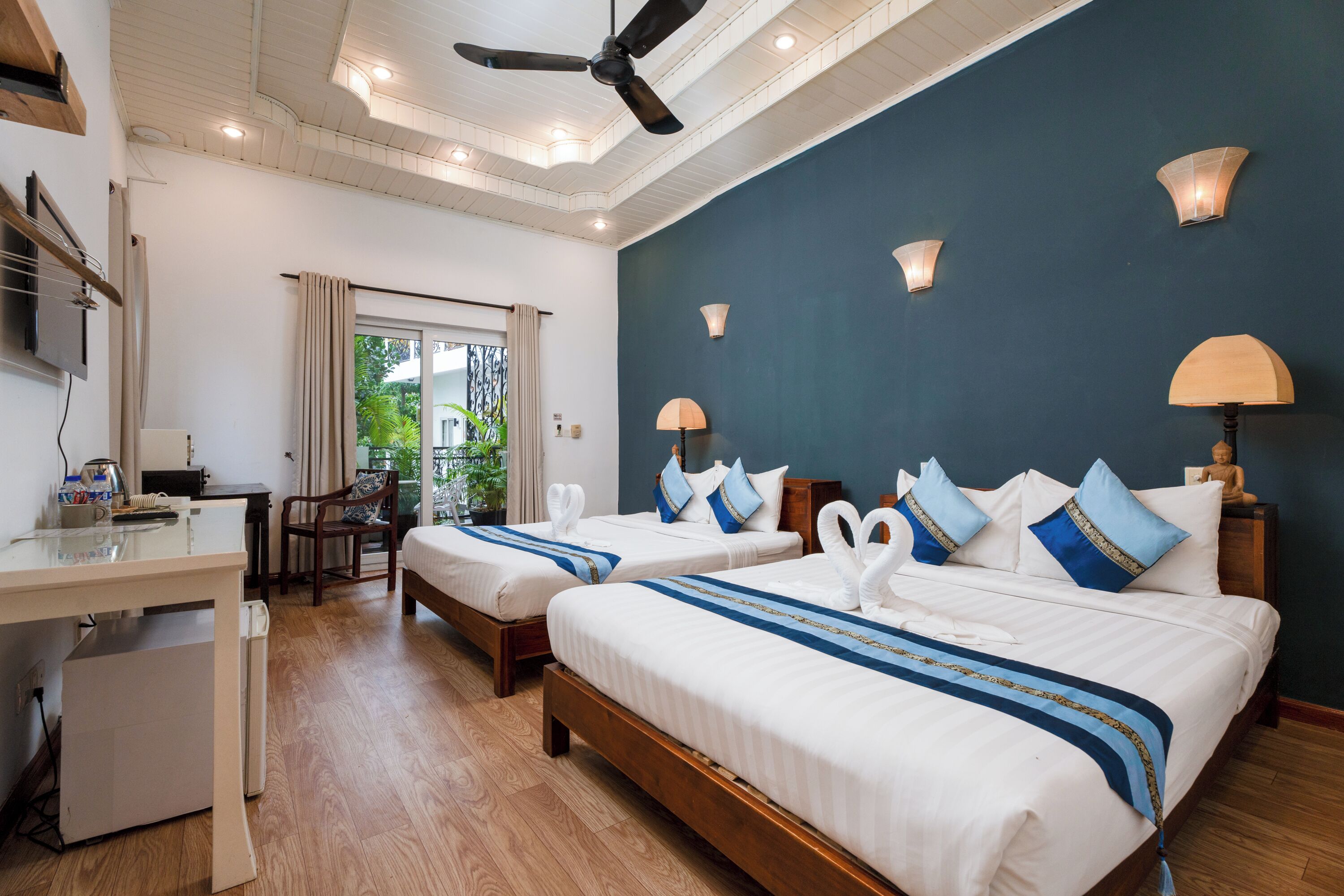 Quarto de hotel com duas camas, uma escrivaninha, uma cadeira e um ventilador de teto.