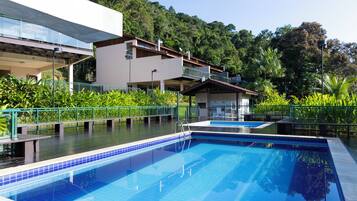 Piscina com borda em azulejos azuis, rodeada por vegetação e um edifício com varanda.