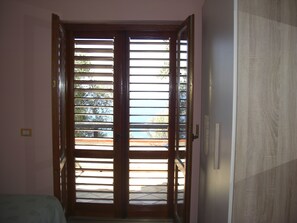 Quarto com porta de madeira, persianas horizontais, cama com colcha estampada e interruptor de luz fixado na parede.