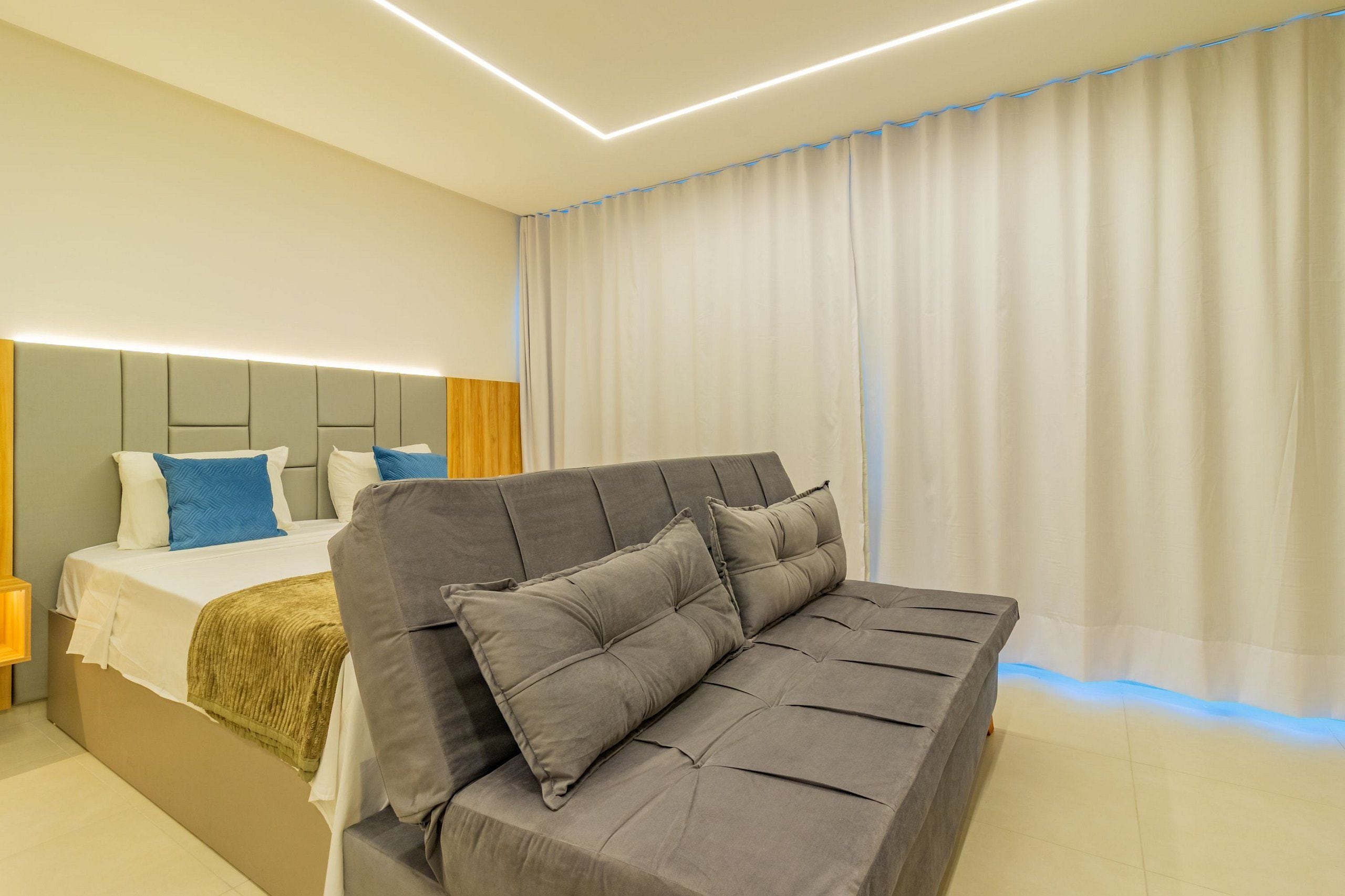Quarto moderno com cama grande, sofá e cabeceira com iluminação embutida.