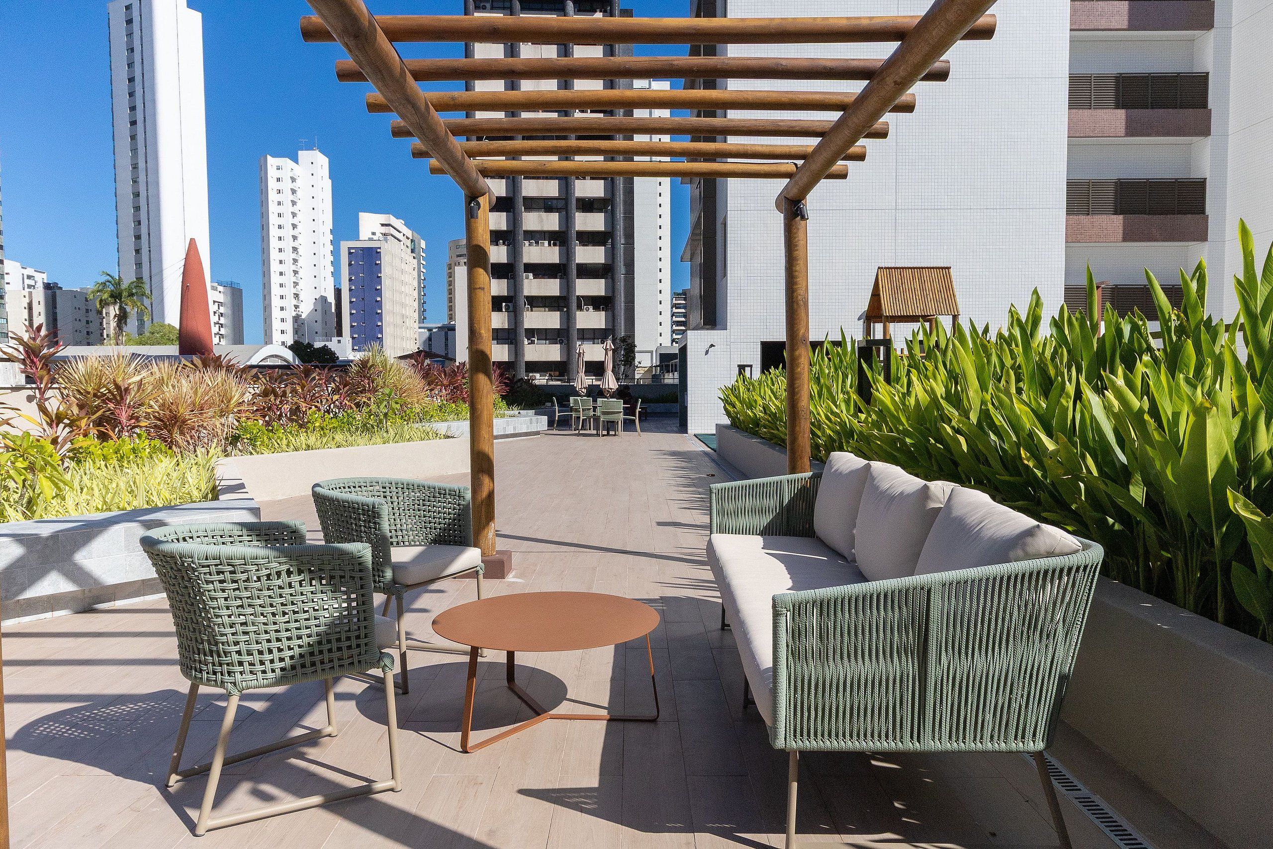 Uma área externa com mobiliário de vime e pérgola, cercada por plantas e vista para a cidade.