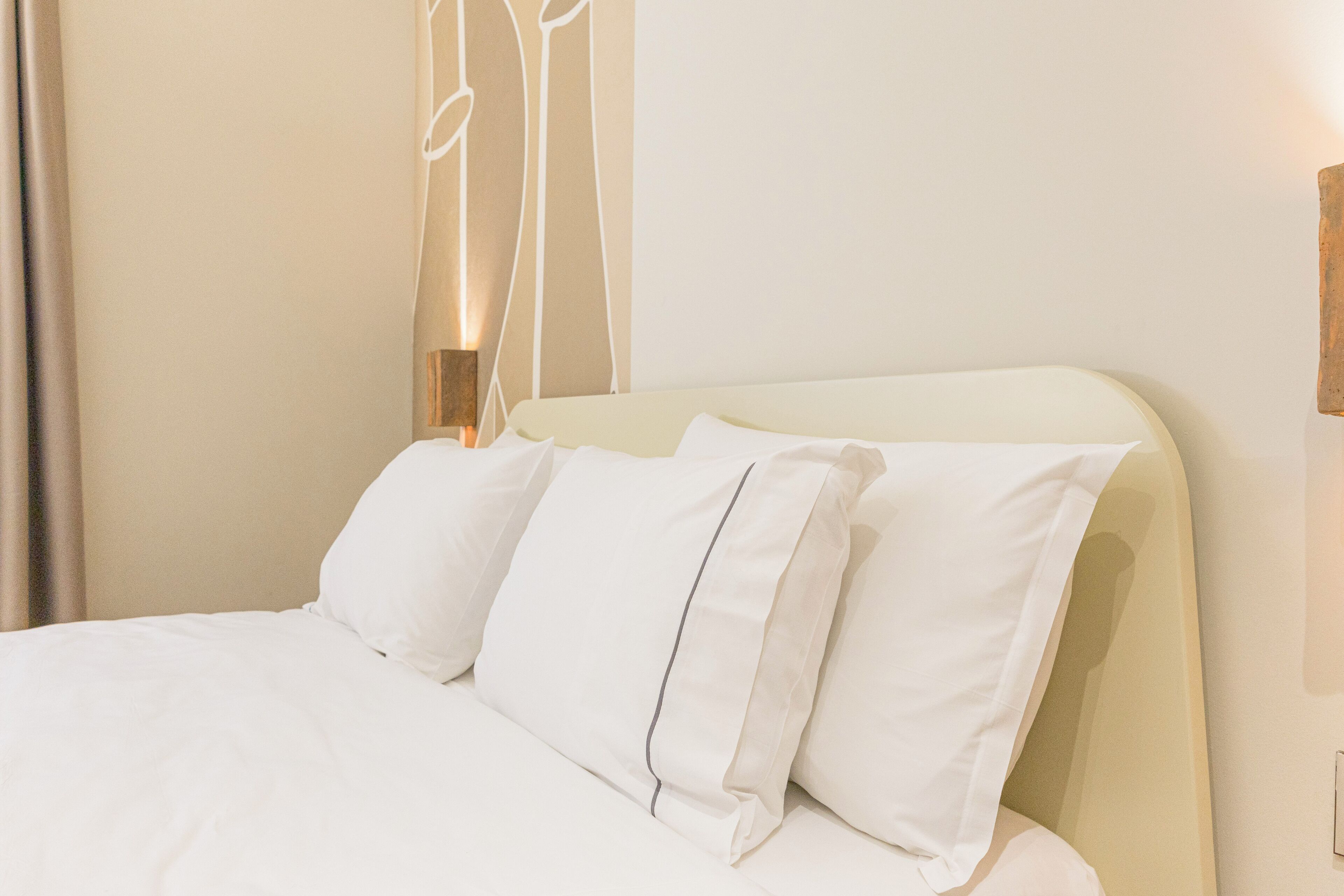 Uma cama bem arrumada com lençóis e almofadas brancos, cabeceira de madeira e luminária fixada na parede.