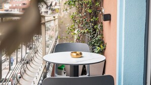 Uma varanda com uma mesa redonda e cadeiras, uma tigela dourada pequena e vista para uma paisagem urbana com edifícios e uma rua.