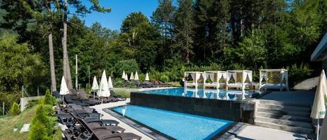 Piscina com espreguiçadeiras e guarda-sóis, cercada por árvores e um edifício com área coberta.