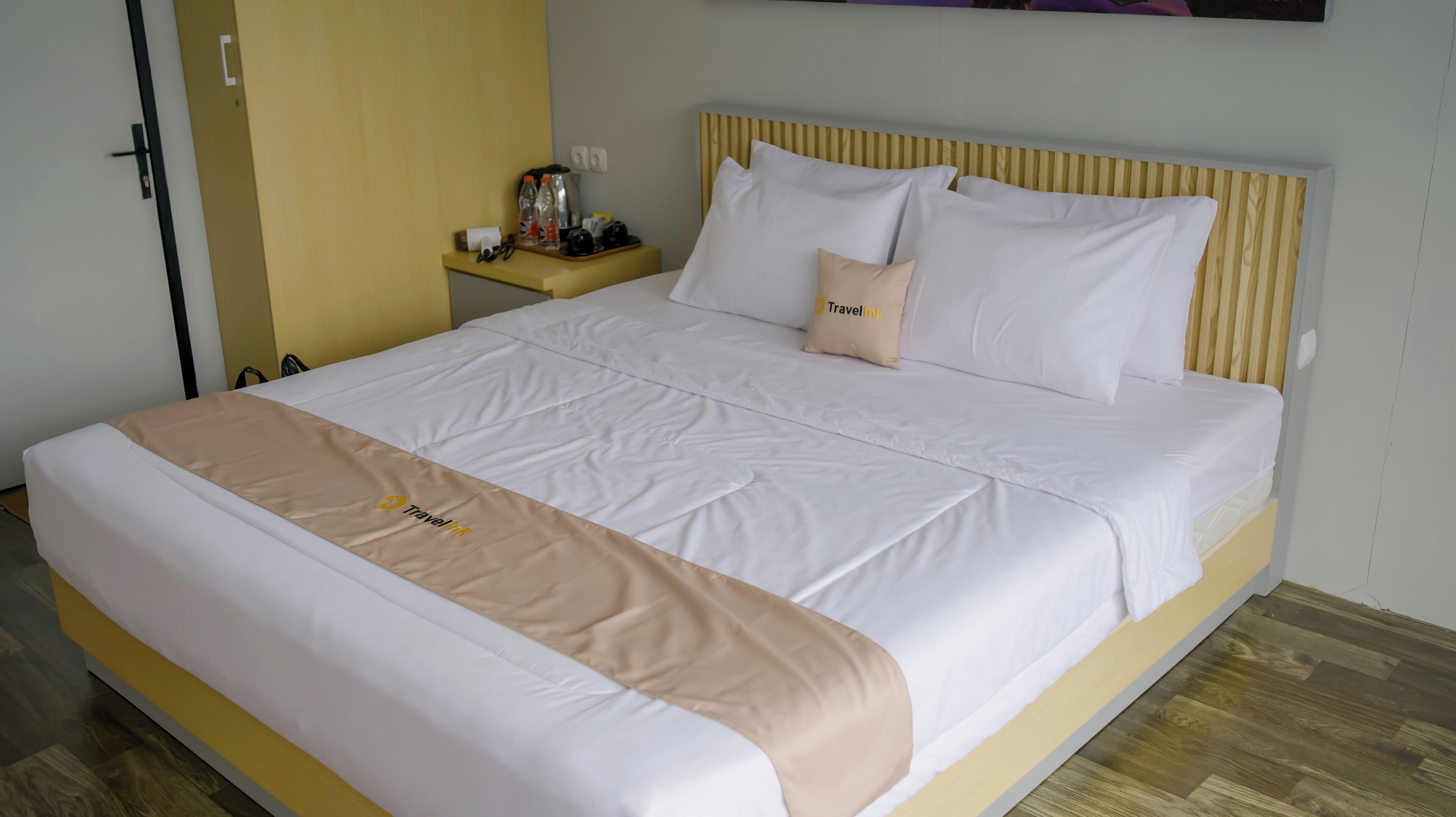 Cama bem arrumada com colcha branca e bege, cabeceira de madeira e mesa de cabeceira com uma garrafa e copos.