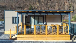 Um edifício moderno com um deck de madeira e guarda-corpo de vidro, apresentando uma escadaria e um espaço de estar com cadeiras.