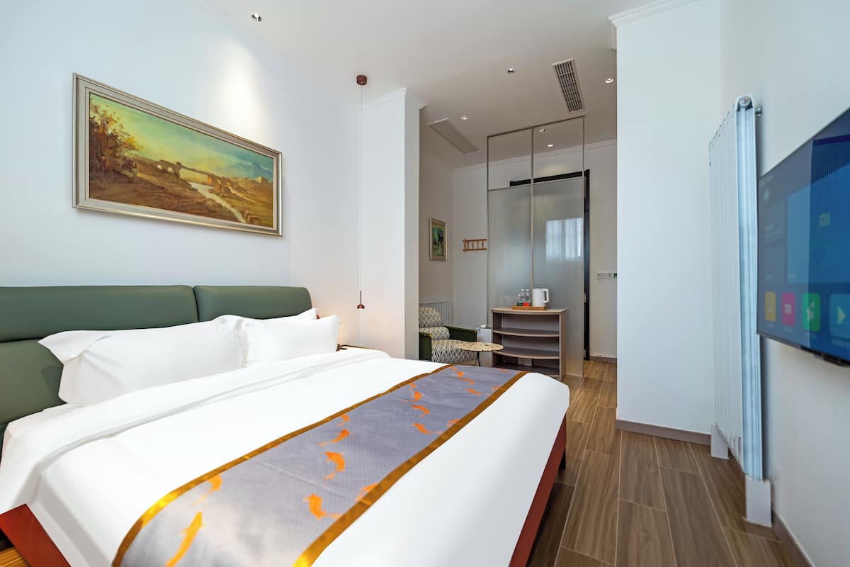 Quarto de hotel com uma cama grande, um quadro na parede, uma televisão e um banheiro ao fundo.